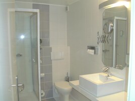 Casa de banho com cabine de duche em vidro, sanita, lavatório com espelho e dispensador de toalhas.
