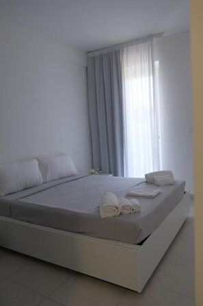 Quarto moderno com cama grande, roupa de cama branca e vista para o exterior através de cortinas leves.