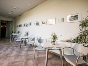 Uma área de recepção com mesa de vidro, cadeiras brancas, plantas em vasos e quadros nas paredes.