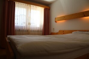Uma cama de solteiro com cabeceira de madeira em quarto com uma janela ampla coberta por cortinas leves.