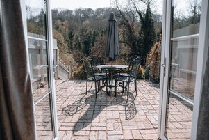 Um terraço ao ar livre com uma mesa redonda e cadeiras, um guarda-sol e vista para uma área arborizada através de uma porta de vidro.
