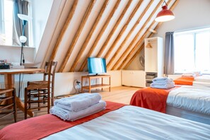 Quarto com teto inclinado, uma escrivaninha, uma cadeira, uma cama com cobertor vermelho e uma janela com cortinas.