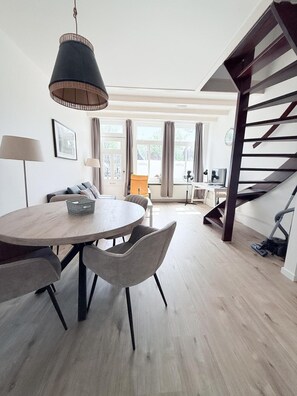 Uma sala de estar moderna com uma mesa de jantar redonda de madeira, cadeiras estofadas em cinza, um lustre, uma escada e vista para o exterior através de amplas janelas.