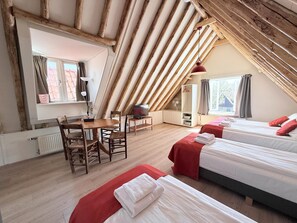 Quarto com teto inclinado, vigas de madeira, mesa de jantar com cadeiras, televisão e duas camas com roupa de cama vermelha.