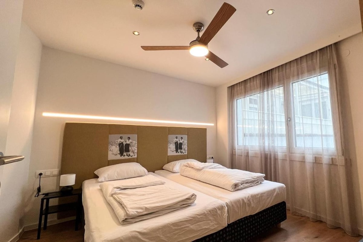 Quarto de hotel com cama, escrivaninha, ventoinha no teto e janela com cortinas.