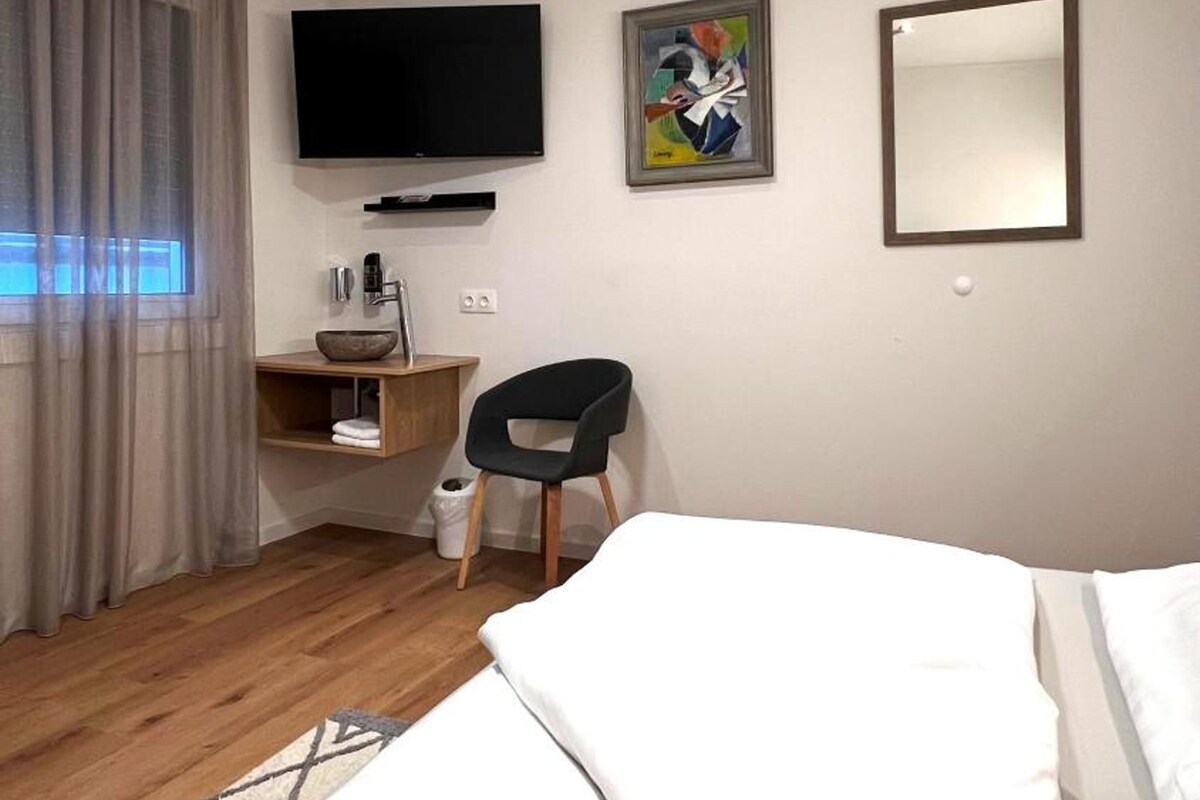 Quarto de hotel com cama, escrivaninha, cadeira, televisão, ventoinha no teto e janela com cortinas.