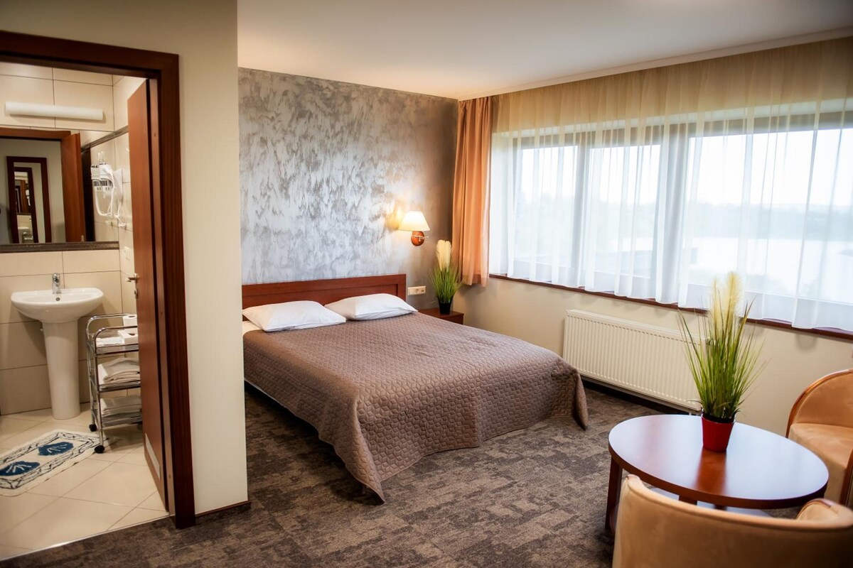 Quarto de hotel com cama, área de estar com poltrona e mesa, banheiro com pia e janela com cortinas.