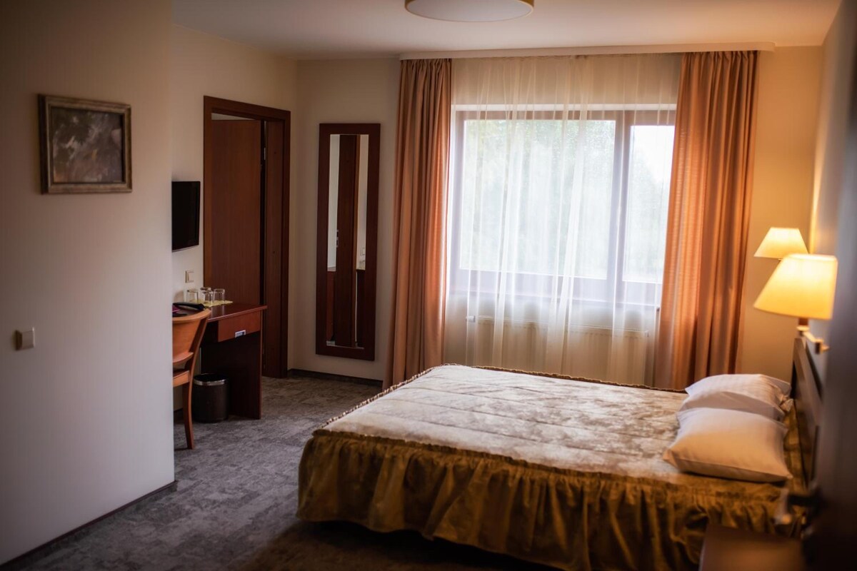 Quarto de hotel com cama, escrivaninha, cadeira, abajur e janela com cortinas.