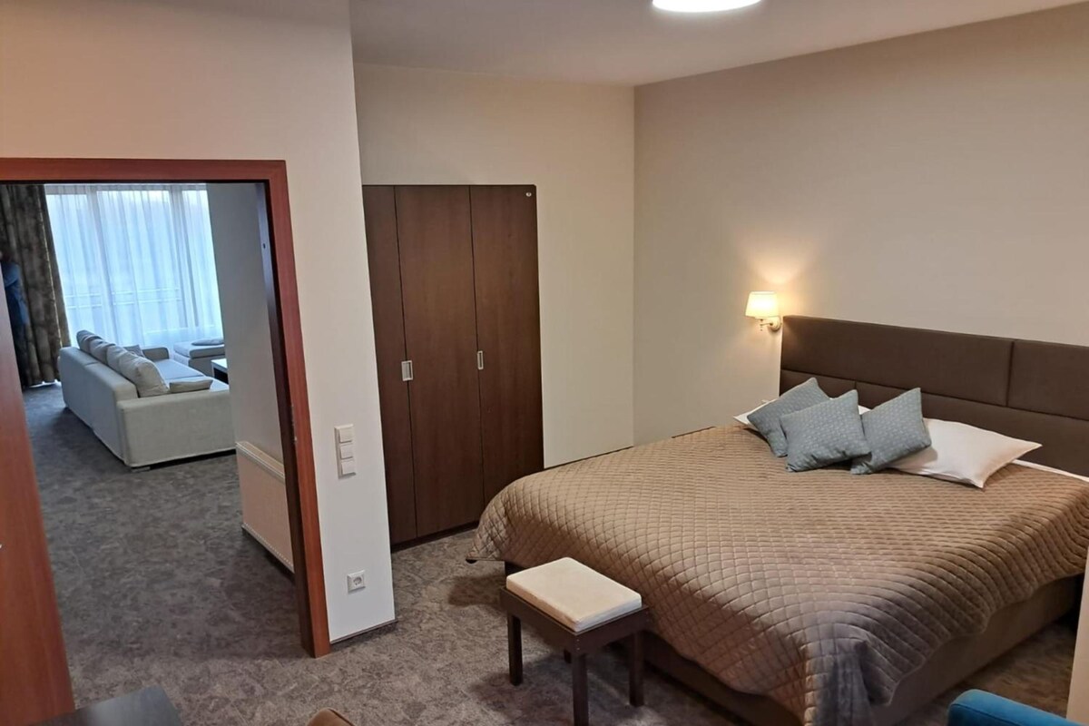 Quarto de hotel com cama, mesa de cabeceira, cadeira e vista para uma sala de estar com sofá e cortinas.