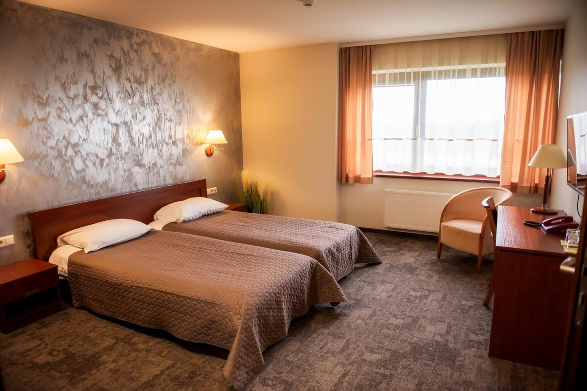 Quarto de hotel com duas camas, uma escrivaninha, uma cadeira, uma janela com cortinas e uma parede com padrão texturizado.