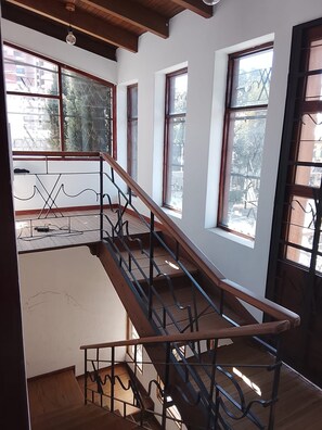 Escadaria com corrimão de metal que leva a um andar superior, janelas amplas com molduras de madeira e vista para o exterior.