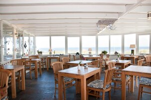 Uma sala de jantar espaçosa com mesas e cadeiras de madeira, grandes janelas com vista para o mar, e um teto com uma escultura decorativa de um pássaro.