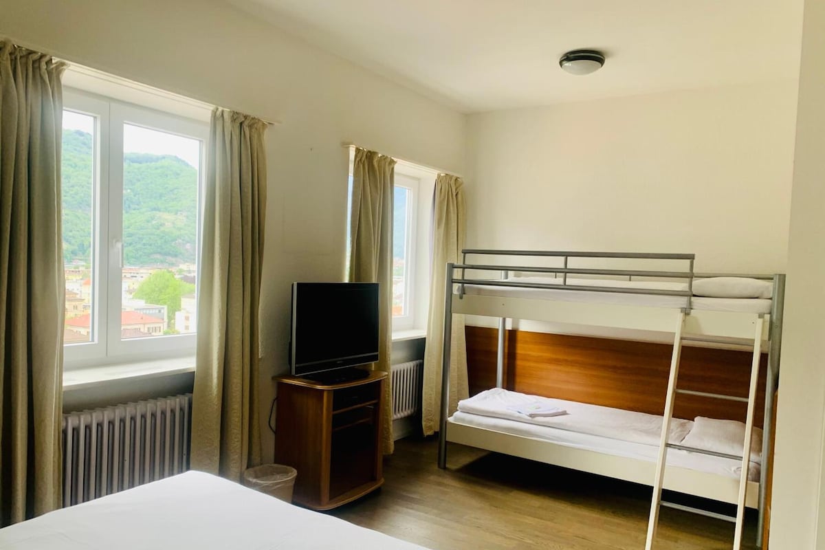 Quarto com beliche, televisão sobre suporte de madeira, radiador e janela com cortinas.