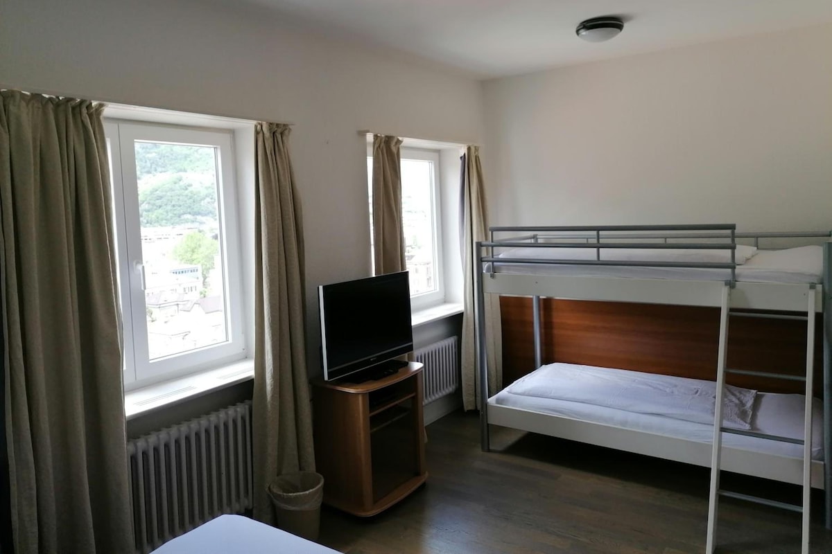 Quarto com beliches, uma televisão sobre um suporte de madeira e janelas com cortinas.