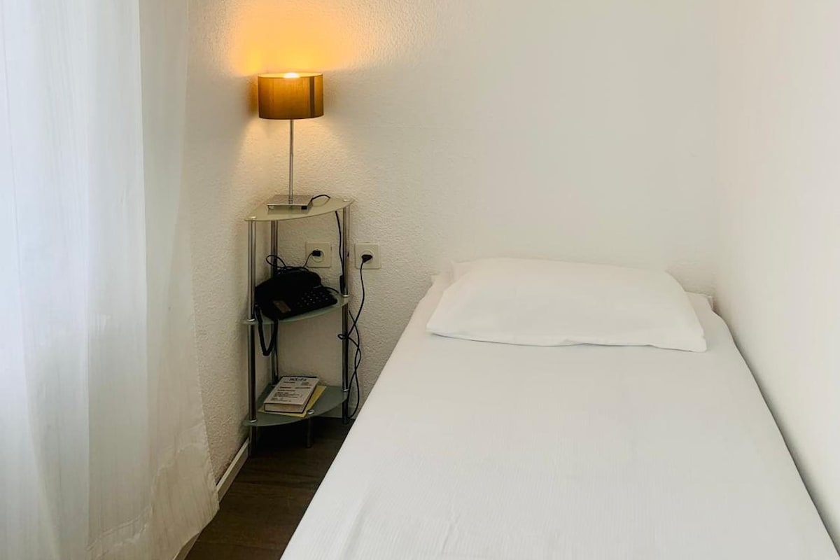 Quarto pequeno com uma cama de solteiro, mesa de cabeceira, luminária e tomada.