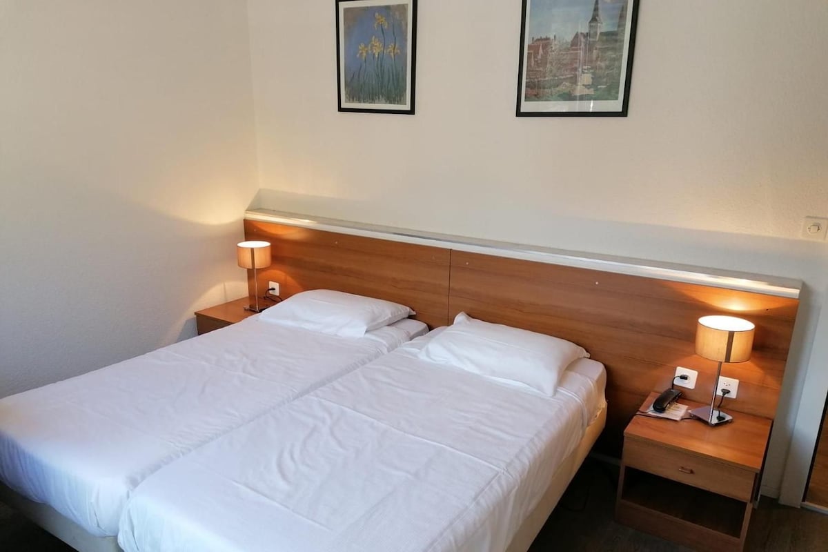 Uma cama de casal com roupa de cama branca, duas mesas de cabeceira com abajures e duas fotografias emolduradas na parede.