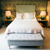 Cama bem arrumada com cabeceira estofada, duas luminárias de cabeceira e um banquinho combinando.