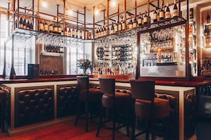 Uma área de bar com várias prateleiras de garrafas e copos, um balcão com uma placa e banquetas de couro.