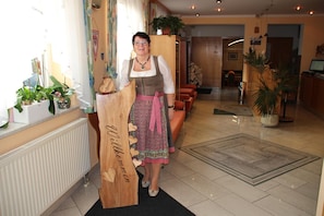 Uma pessoa vestida com traje tradicional segura uma placa de madeira com a palavra 'Willkommen', em um saguão de hotel com radiador, plantas em vasos e uma área de estar ao fundo.