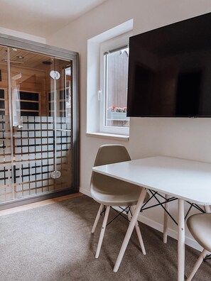 Quarto com mesa branca, duas cadeiras, porta de vidro deslizante para um interior de madeira, janela com vista para flores e TV de tela plana montada na parede.