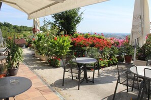 Um pátio externo com mesas e cadeiras, rodeado por vasos de plantas e flores.