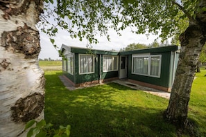 Uma casa móvel verde com duas janelas e uma porta, rodeada por relva e árvores.