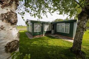 Uma casa móvel verde com duas janelas e uma porta, rodeada por relva e árvores.