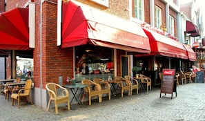 Edifício de tijolos com área externa para sentar e toldos vermelhos, exibindo uma placa de restaurante e um anúncio da Coca-Cola.
