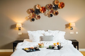 Quarto de hotel com cama, almofadas, mesas de cabeceira, luminárias na parede e bandeja com itens de café da manhã.