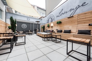 Área de estar externa com bancos e mesas de madeira, uma grande parede com a palavra 'Lounge' e um logotipo, e piso ladrilhado.