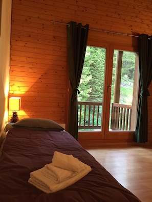 Quarto com painéis de madeira, uma cama, mesa de cabeceira, luminária e vista para uma varanda com cortinas.