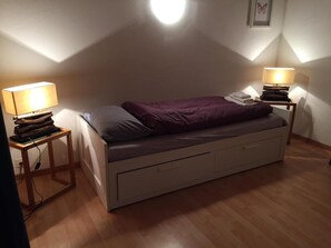 Quarto individual com uma cama, piso de madeira, duas mesas de cabeceira com luminárias, uma luminária na parede e um quadro emoldurado.