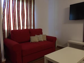 Um sofá vermelho com duas almofadas, uma mesa de centro branca e uma TV fixada na parede.