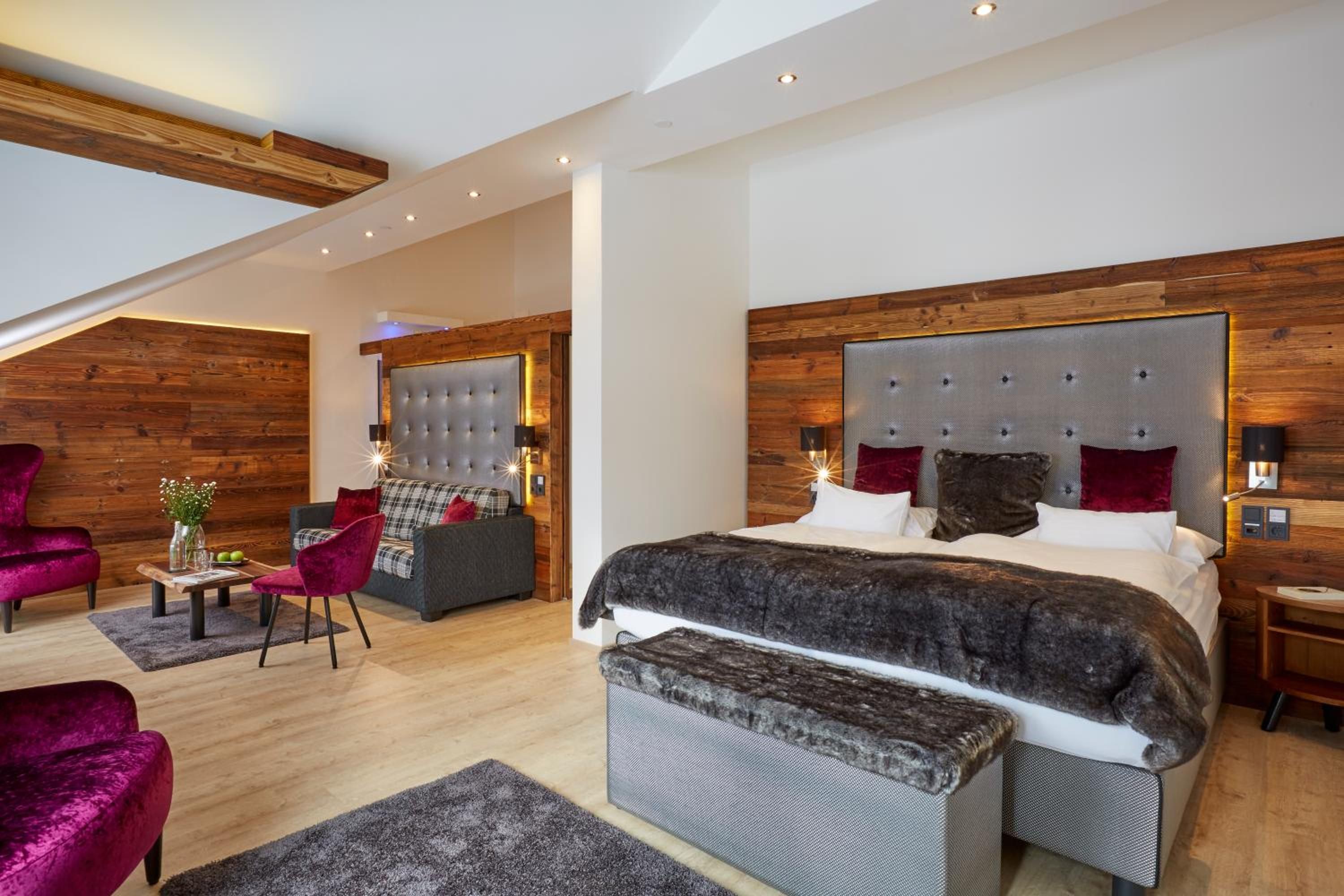Quarto moderno com cama grande, área de estar com sofá e cadeiras, e teto de madeira.
