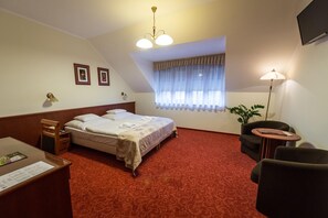 Quarto de hotel com cama, escrivaninha, cadeira, planta e janela com cortinas.