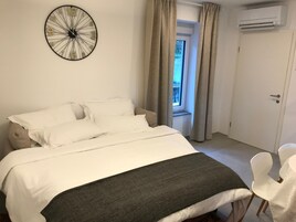 Um quarto com cama, relógio na parede, janela com cortinas e ar condicionado.