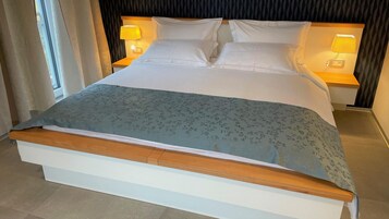 Quarto de hotel com cama, duas luminárias laterais, janela com cortinas e cabeceira de madeira.