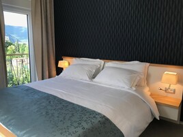 Quarto de hotel com cama, duas luminárias de cabeceira, vista para vegetação e montanhas através de uma janela e uma parede texturizada escura.