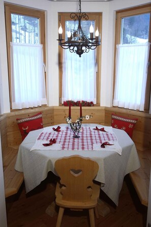 Uma sala de jantar com mesa posta para dois, um lustre e um banco com almofadas vermelhas.