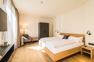 Quarto de hotel com cama, mesas de cabeceira, escrivaninha, cadeira, guarda-roupa e televisão.
