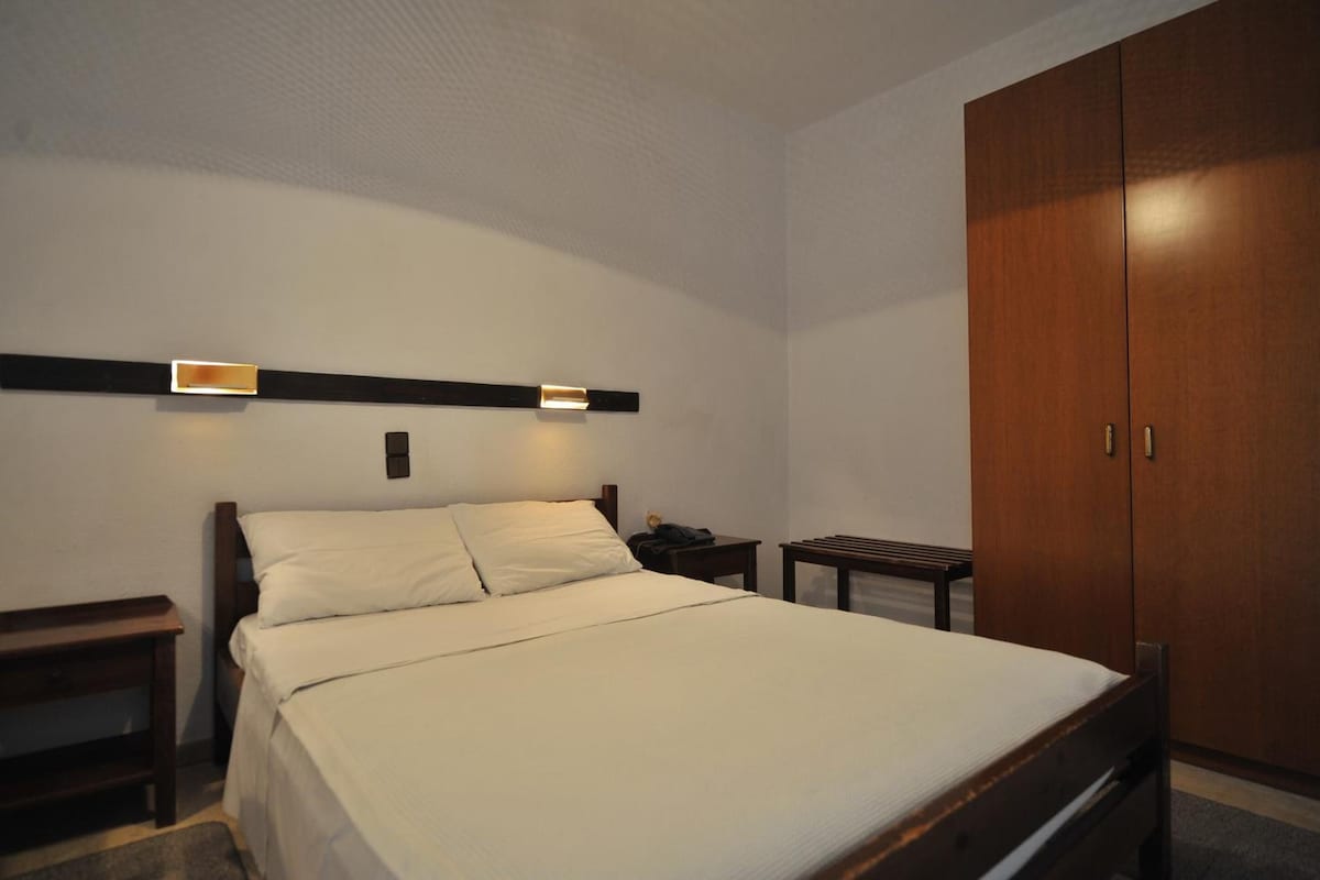 Quarto com cama, mesa de cabeceira, guarda-roupa e luminária na parede.
