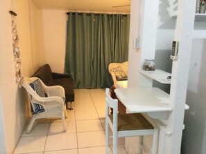 Um quarto pequeno com uma cadeira branca, um sofá castanho, uma cortina verde e uma mesa branca.