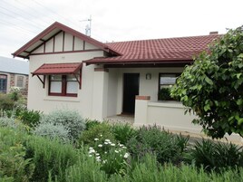 Uma casa de dois andares com telhado de telhas vermelhas, varanda frontal e jardim com diversas plantas e flores.