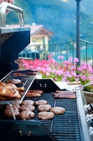 Uma churrasqueira com diversos tipos de carne sendo preparados ao ar livre, com montanhas e flores cor-de-rosa ao fundo.
