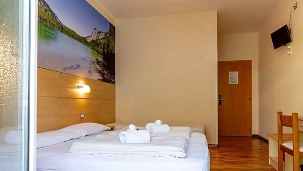 Quarto de hotel com uma cama, cabeceira de madeira, pintura de uma paisagem, televisão na parede e uma cadeira de madeira.