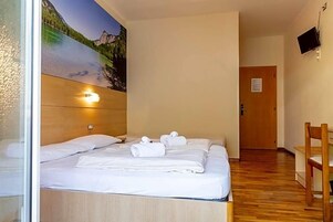 Quarto de hotel com uma cama, cabeceira de madeira, pintura de uma paisagem, televisão na parede e uma cadeira de madeira.