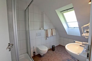Casa de banho pequena e moderna com chuveiro, sanita e lavatório, apresentando um claraboia e paredes revestidas a azulejos.