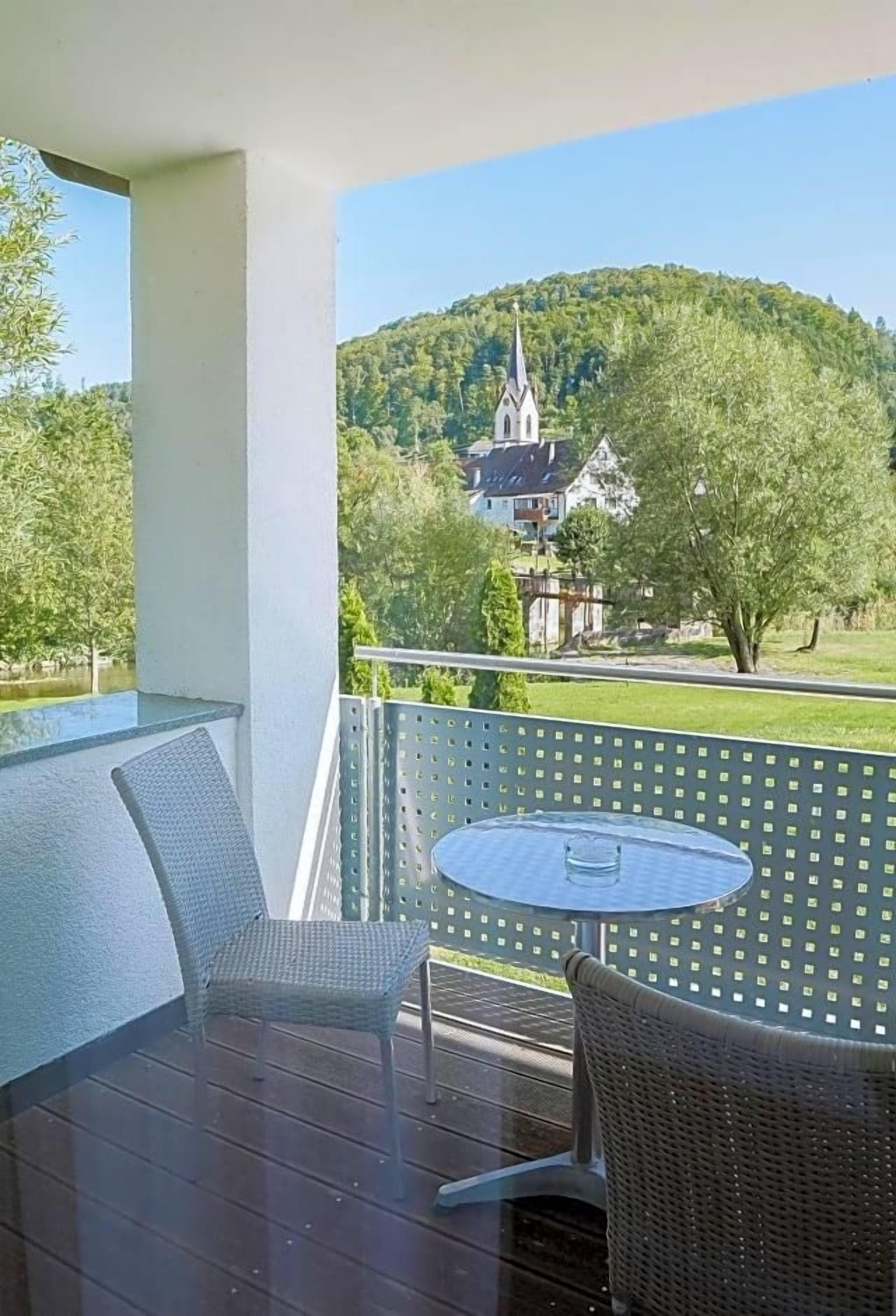 Varanda com mesa e duas cadeiras, dando vista para uma paisagem verde com uma igreja e casas.