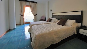 Quarto de hotel com duas camas, mesa de cabeceira, janela com cortinas e chão de azulejos.