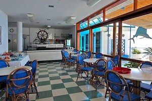Uma sala de jantar com piso xadrez, cadeiras azuis e uma ilha central decorada com um leme de navio.